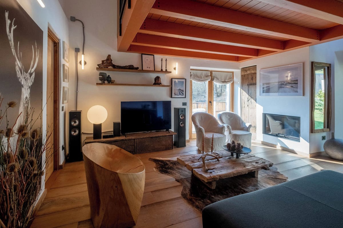 Uma sala de estar moderna com teto de madeira, televisão de tela plana, cadeiras brancas, mesa de centro e lareira.