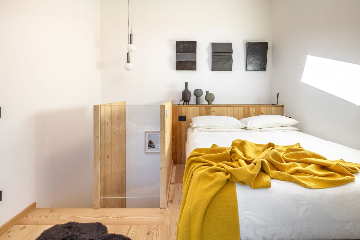 Quarto moderno com cabeceira de madeira, cama com roupa de cama branca e manta amarela, e uma pequena mesa de cabeceira com objetos decorativos.