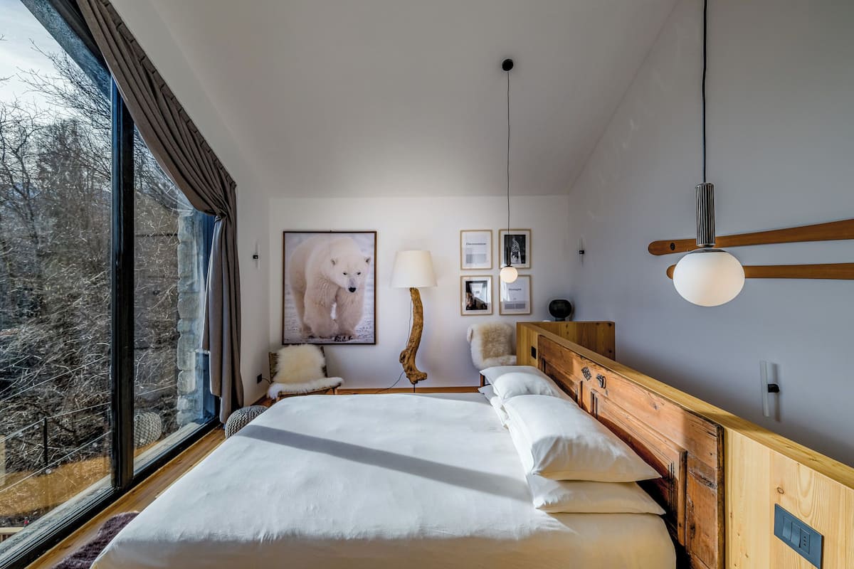Quarto moderno com cama grande, cabeceira de madeira, janela com vista para árvores e luminária fixada na parede.