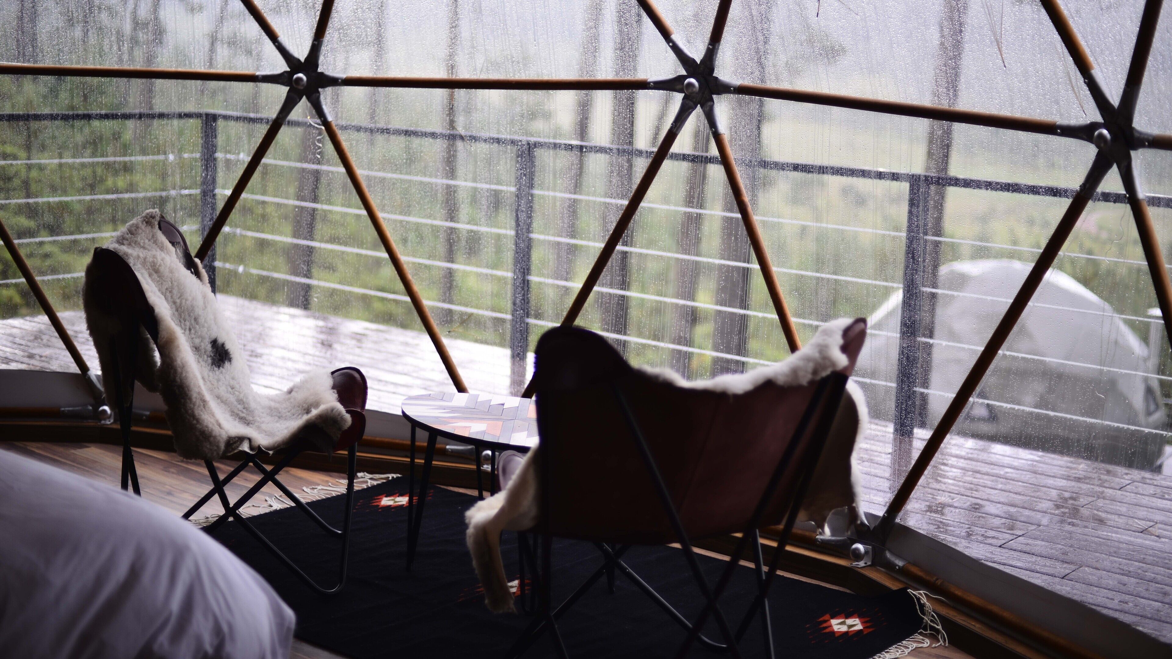 Estrutura em cúpula geodésica com vista para uma área externa chuvosa. No interior, dois cadeiras e uma pequena mesa.