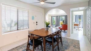 Uma sala de jantar com mesa de madeira, cadeiras e um ventilador de teto. Há uma janela com persianas, uma porta e vista para outro cômodo.