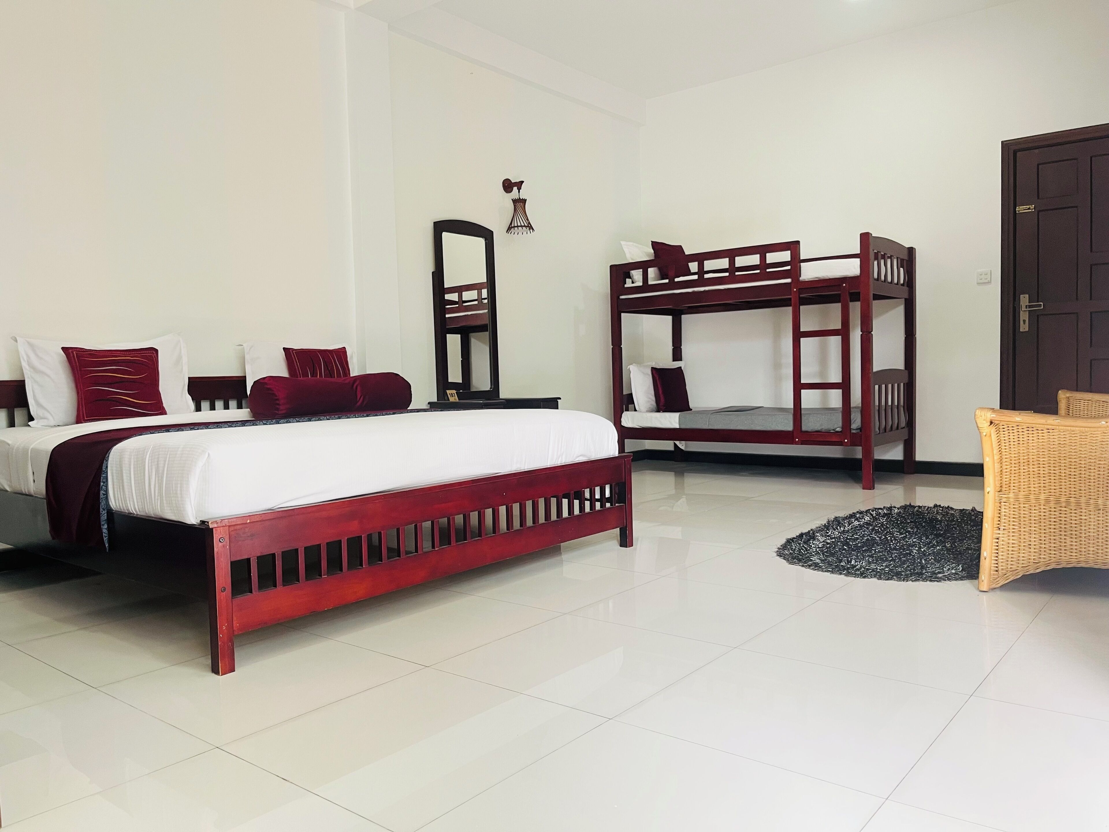 Quarto de hotel com uma cama grande, uma cama menor, um espelho, uma porta e uma cadeira de vime.