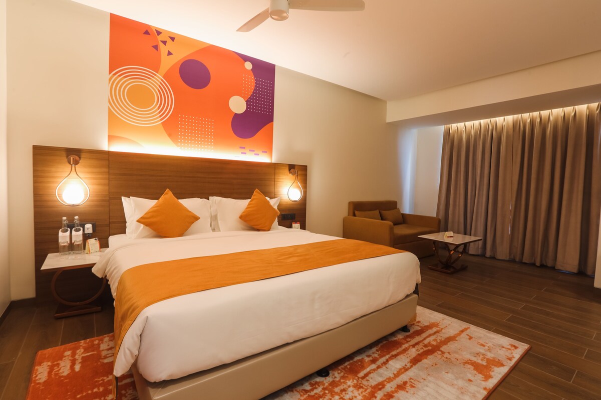 Quarto de hotel com uma cama grande, duas luminárias de cabeceira, cabeceira com luminária embutida, uma mesinha, uma cadeira e uma janela ampla com cortinas.