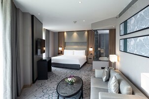Quarto de hotel moderno com uma cama grande, área de estar com sofá e mesa de centro, televisão e banheiro com espelho e pia.