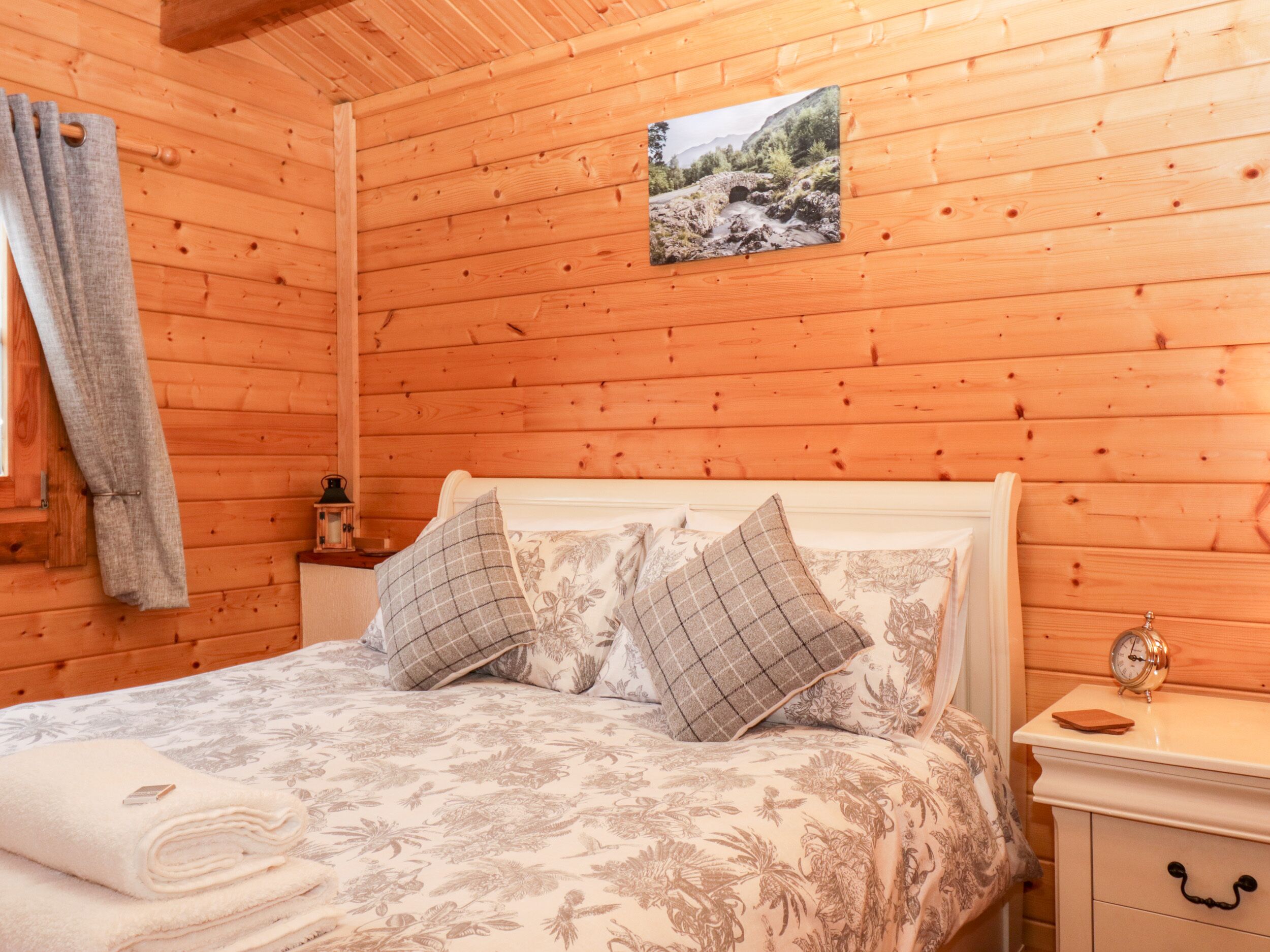 Quarto de cabana de madeira com cama, mesa de cabeceira, quadro na parede e janela com cortinas.