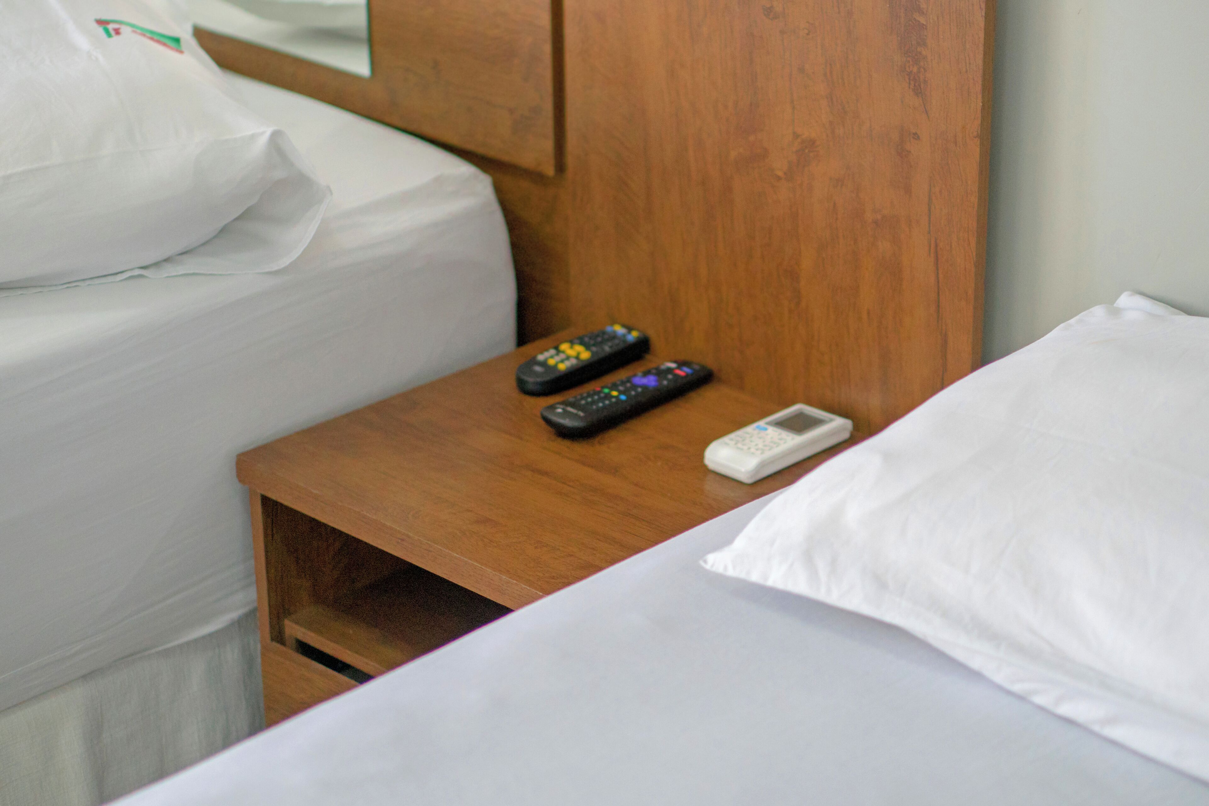 Uma cama bem arrumada com lençóis brancos, uma mesinha de cabeceira de madeira com dois controles remotos e um travesseiro branco.