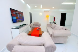 Uma sala de estar moderna com uma TV de tela plana na parede, uma mesa de centro vermelha, dois sofás bege e uma pequena mesinha lateral.