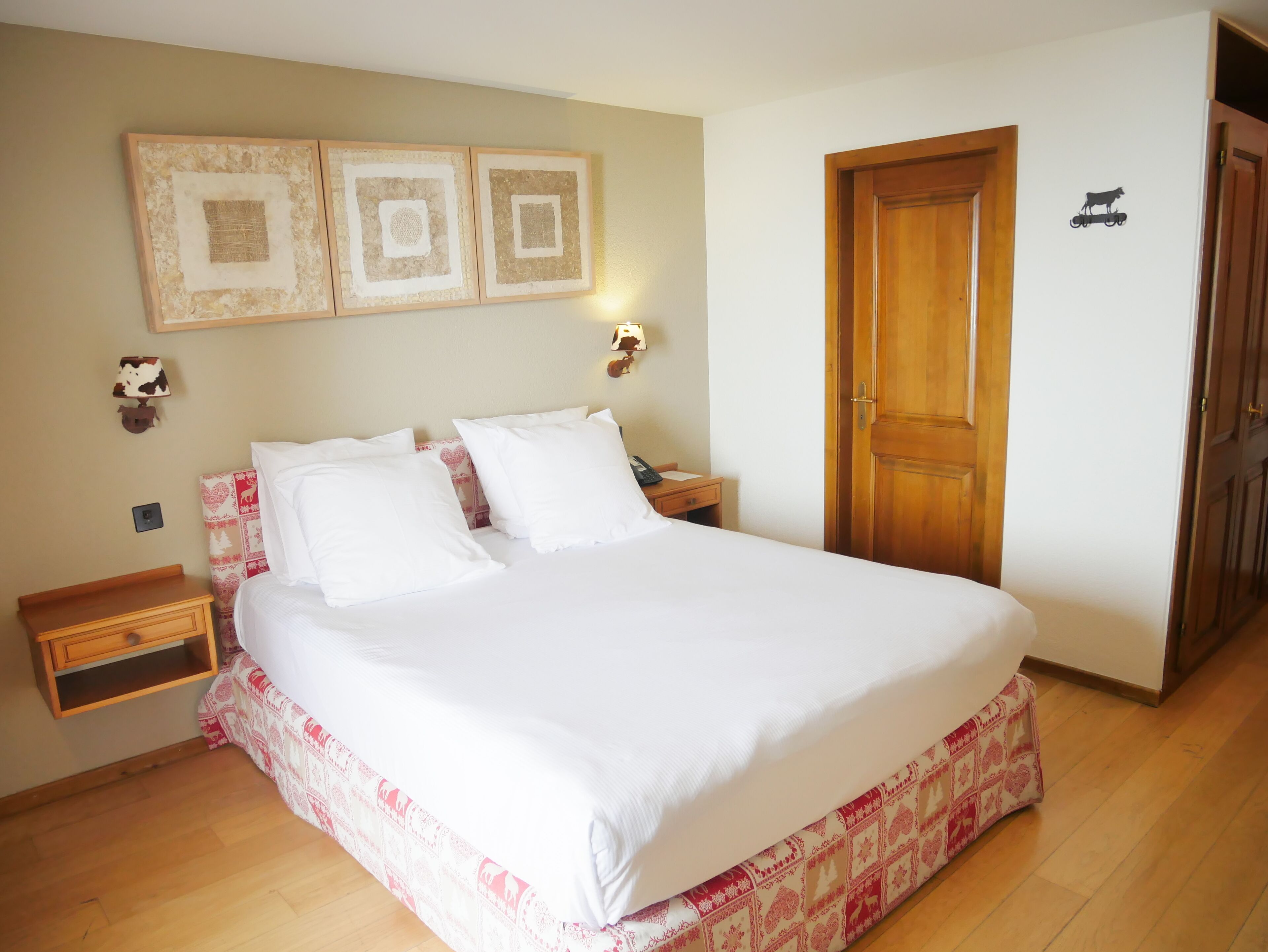 Cama bem arrumada com lençóis brancos, cabeceira de madeira e saia de cama estampada. Há duas mesas de cabeceira com luminárias, uma porta de madeira e um luminário fixado na parede.