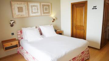 Cama bem arrumada com lençóis brancos, cabeceira de madeira e saia de cama estampada. Há duas mesas de cabeceira com luminárias, uma porta de madeira e um luminário fixado na parede.