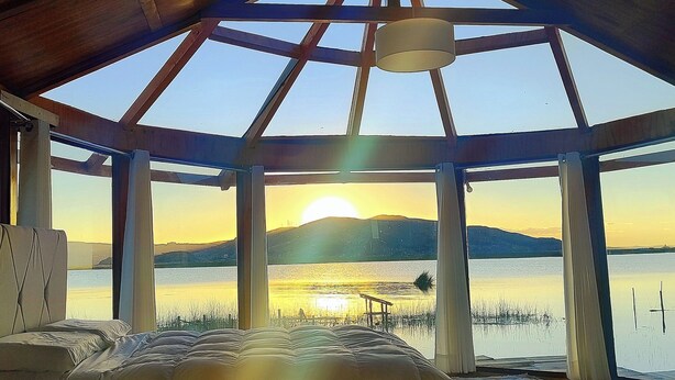 Quarto com um grande claraboia, cama e vista para um lago e montanhas ao pôr do sol.