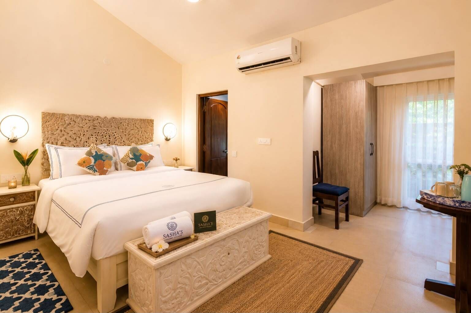 Quarto de hotel com cama, mesas de cabeceira, ar condicionado, uma cadeira, uma pequena mesa com um vaso e uma porta que dá para outro cômodo.