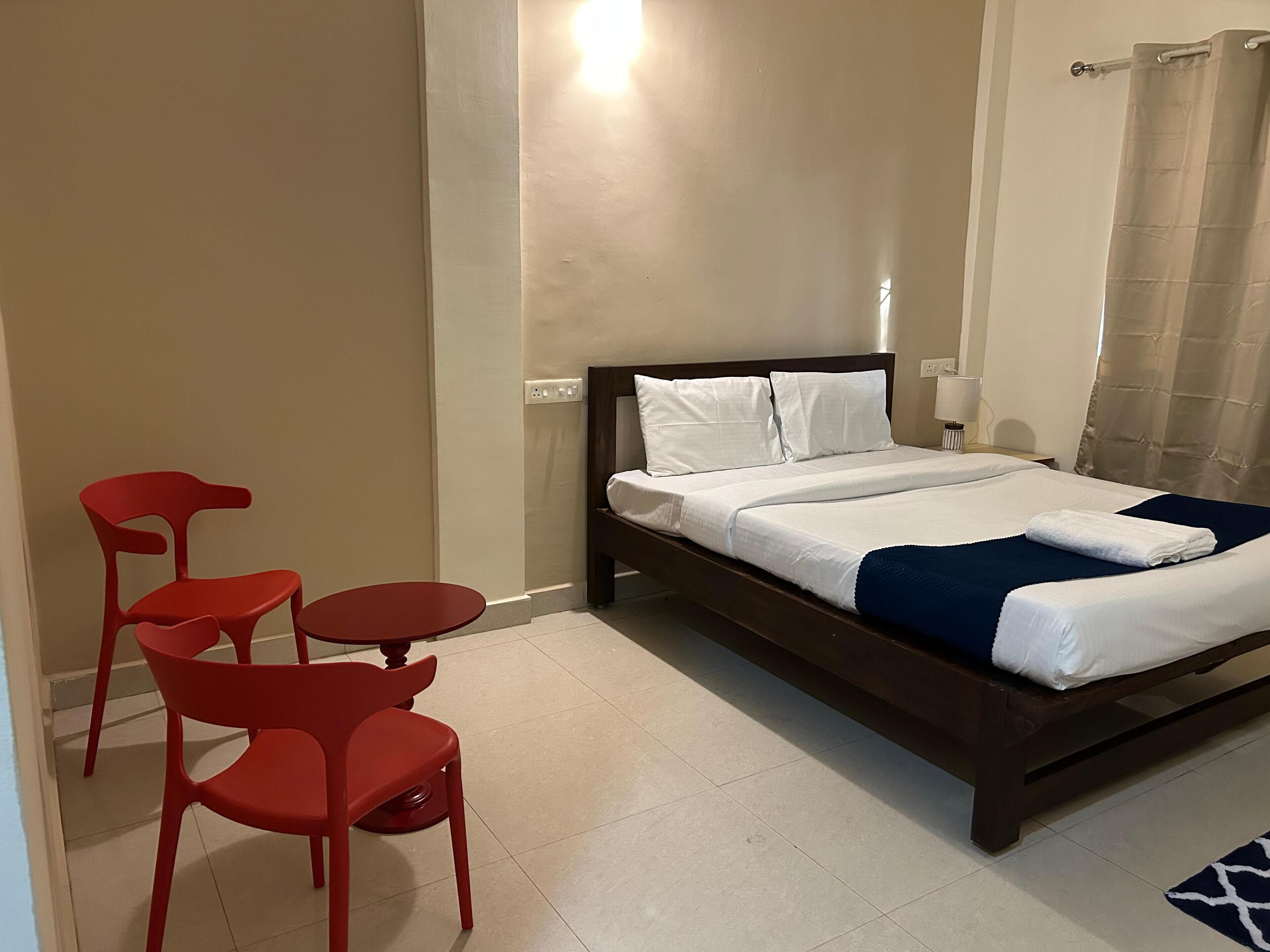 Quarto com cama, duas cadeiras vermelhas, uma mesa redonda pequena e um criado-mudo com abajur.