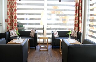 Quarto com dois sofás pretos, uma pequena mesa de madeira e vista pela janela ampla com cortinas estampadas.