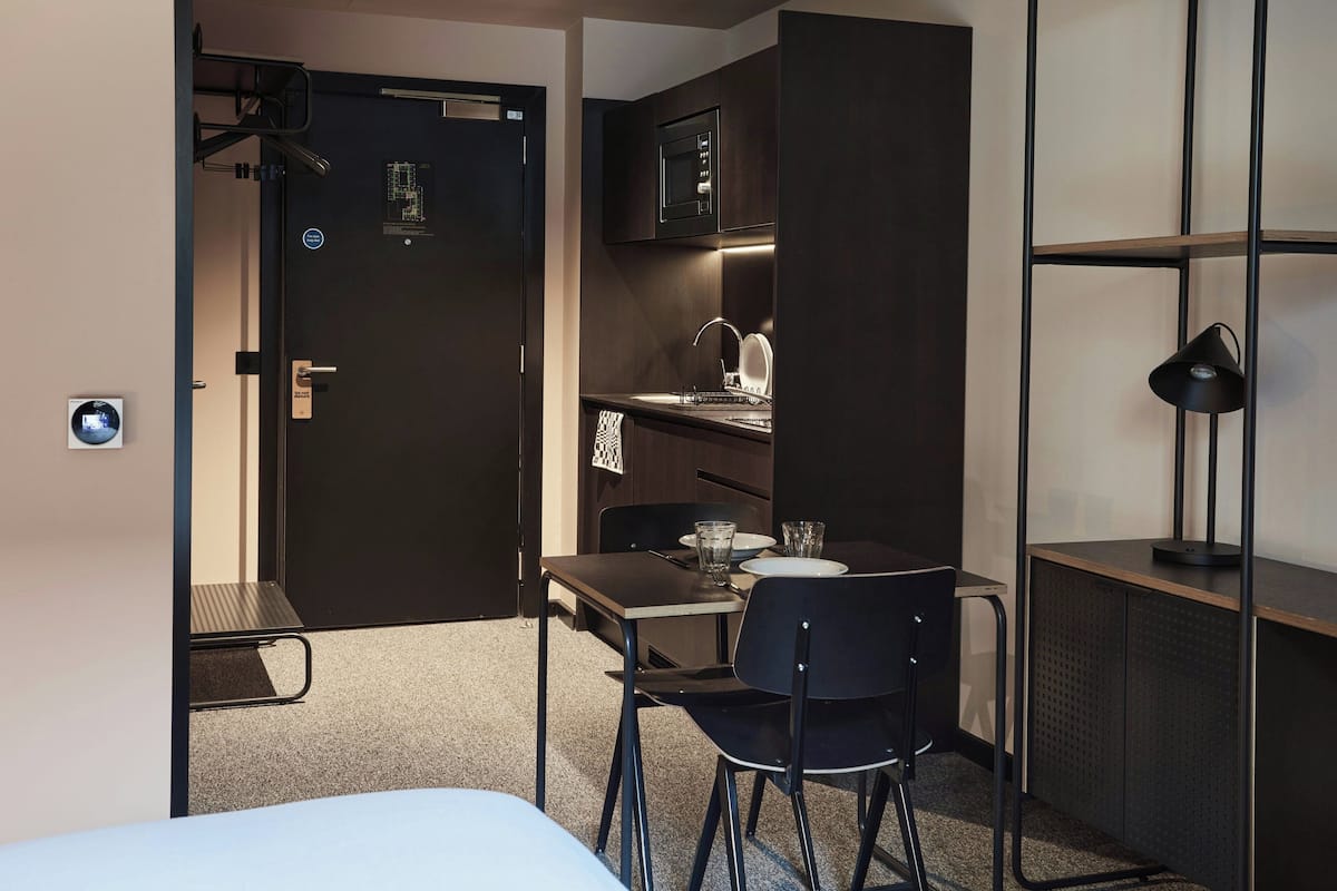 Quarto de hotel compacto com cama, uma pequena mesa com cadeira, cozinha compacta e porta com câmera de segurança.