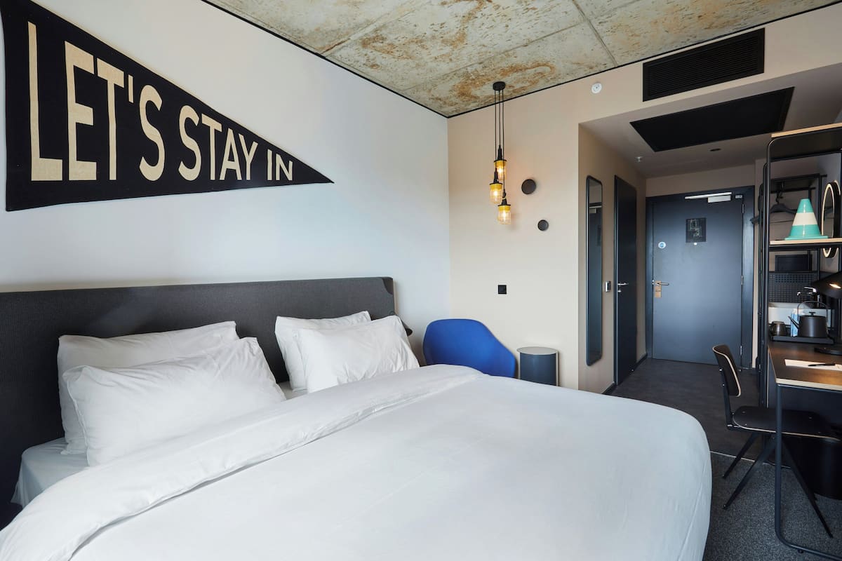 Quarto de hotel com cama, um letreiro 'LET'S STAY IN', escrivaninha com luminária e cozinha compacta.