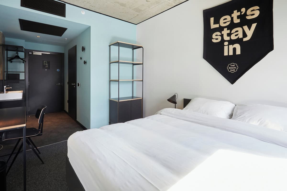 Quarto de hotel moderno com cama, escrivaninha, cadeira, guarda-roupa e prateleira na parede.