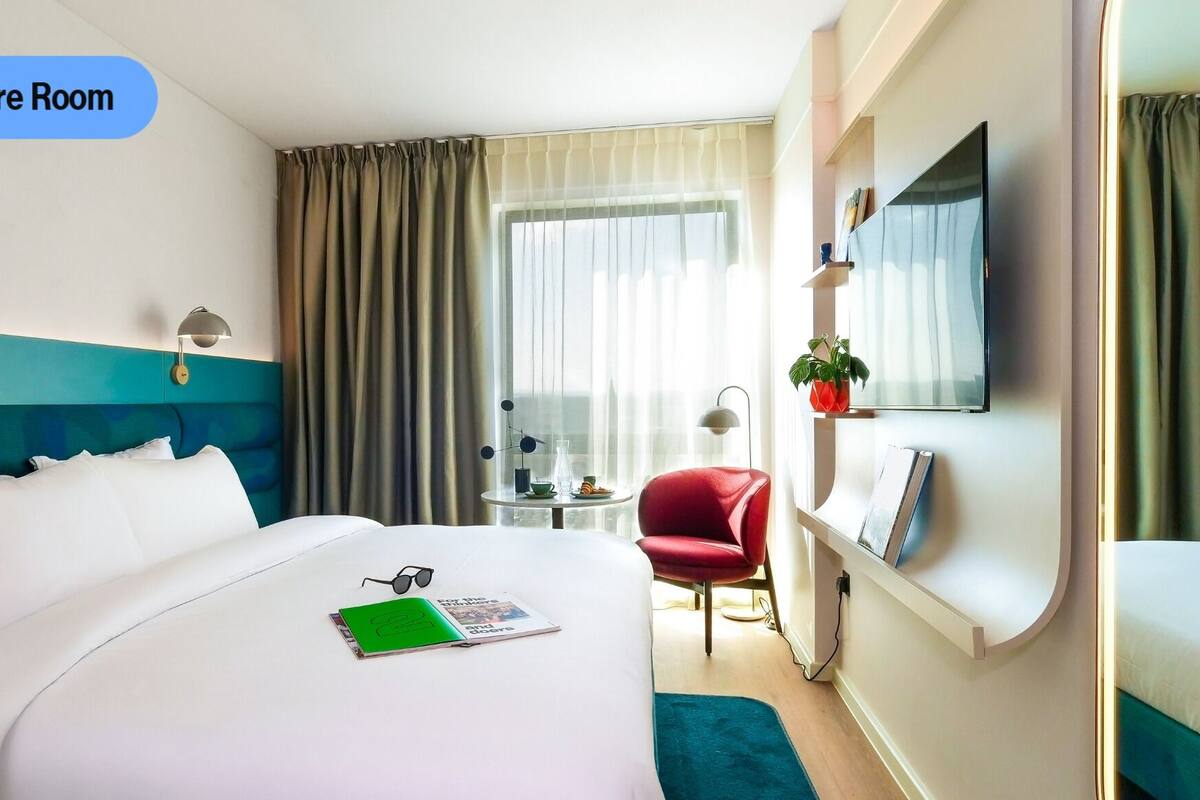 Quarto de hotel com cama, escrivaninha, cadeira, televisão e janela com cortinas.