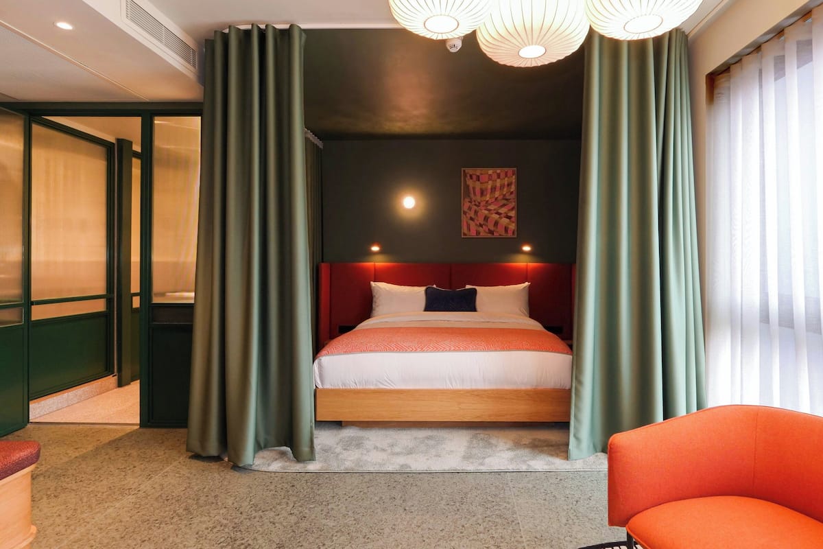 Quarto de hotel com cama, cortinas verdes, cabeceira vermelha, um quadro na parede e uma luminária de teto clara.