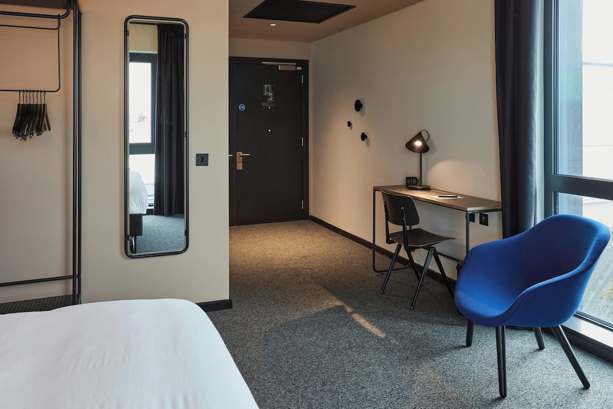 Quarto de hotel com cama, escrivaninha, cadeira azul, espelho e porta.