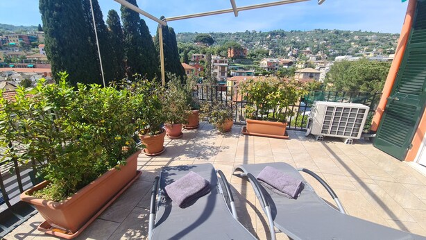 Terraço no telhado com espreguiçadeiras, plantas em vasos e vista para uma vila na encosta.