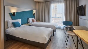 Quarto de hotel com duas camas, uma escrivaninha, uma cadeira, televisão e uma janela grande com cortinas.