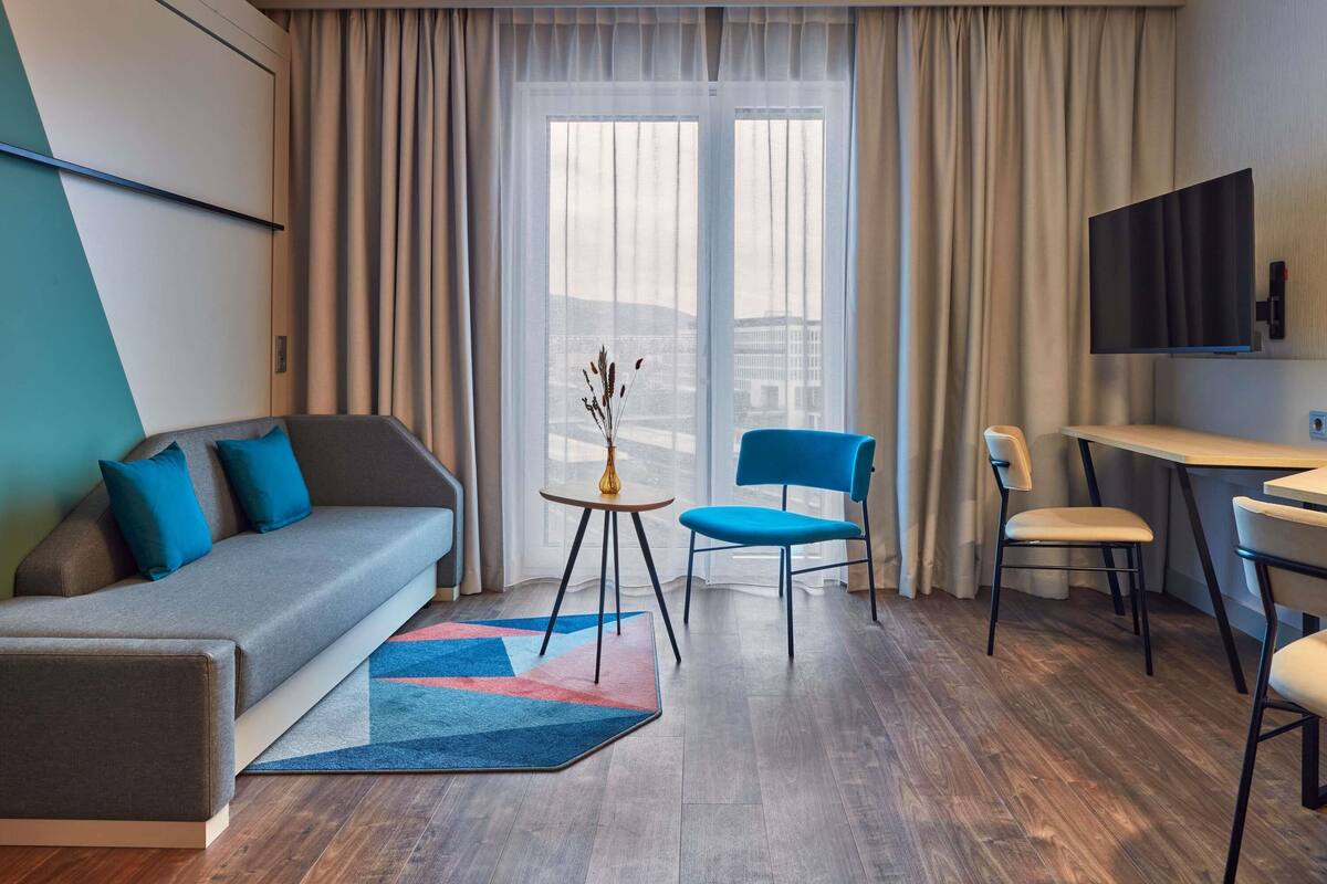 Quarto de hotel moderno com sofá, cadeiras, uma mesa pequena e televisão.