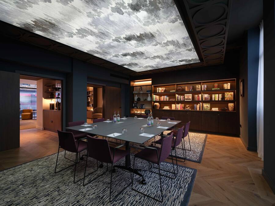Uma sala de reunião moderna com uma mesa grande, cadeiras e um teto com design que lembra nuvens. Há uma estante com livros e objetos decorativos.