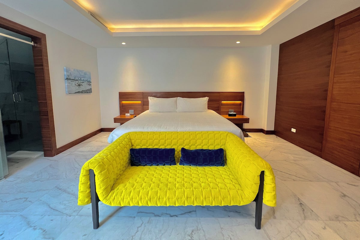 Quarto de hotel moderno com uma cama grande, uma poltrona amarela vibrante, cabeceira de madeira e piso de mármore.