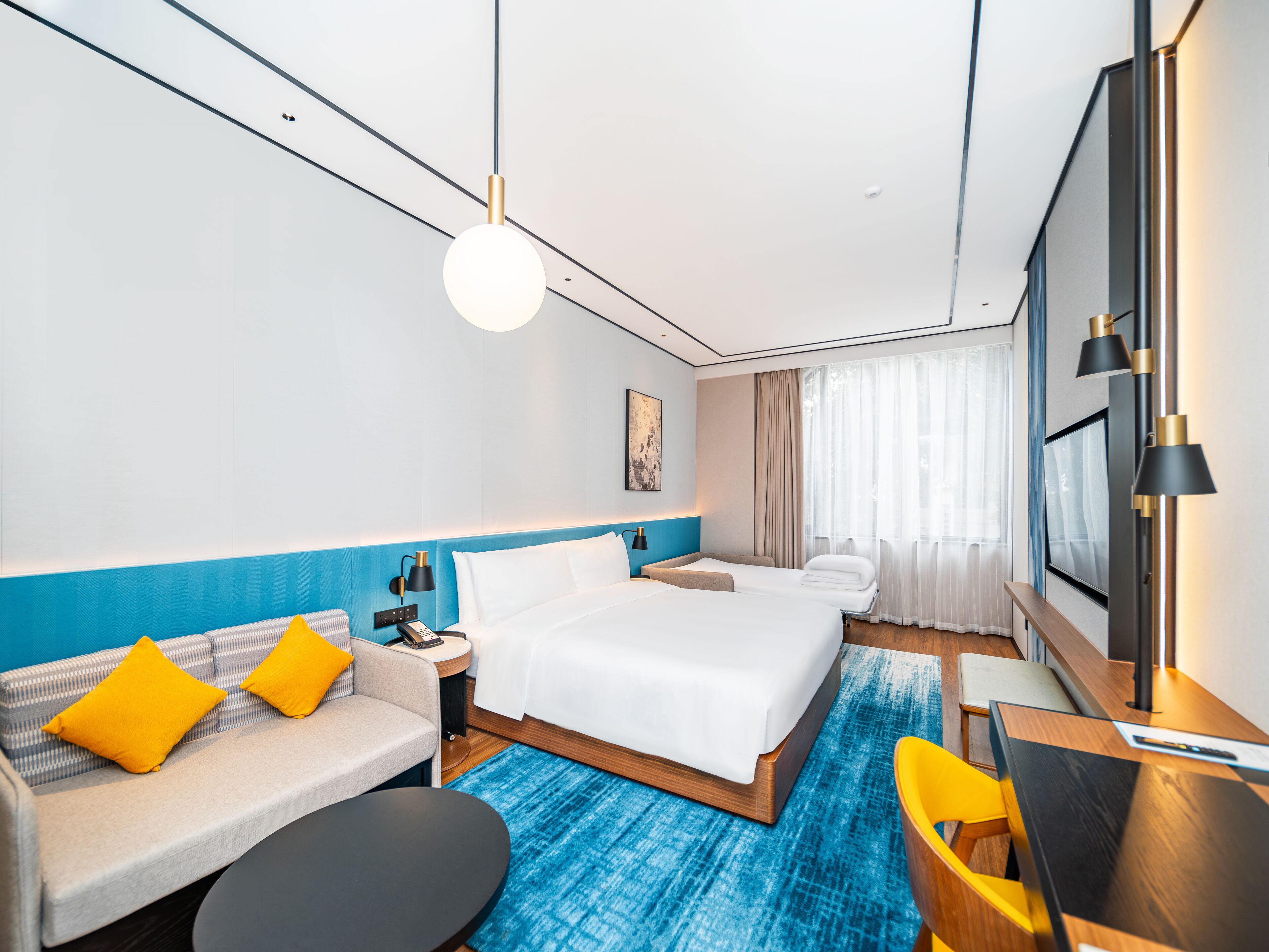 Quarto de hotel com cama, sofá com almofadas amarelas, escrivaninha e televisão.