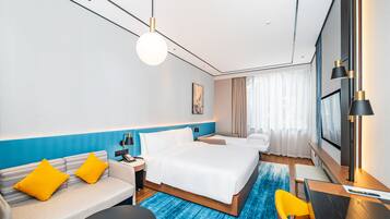 Quarto de hotel com cama, sofá com almofadas amarelas, escrivaninha e televisão.