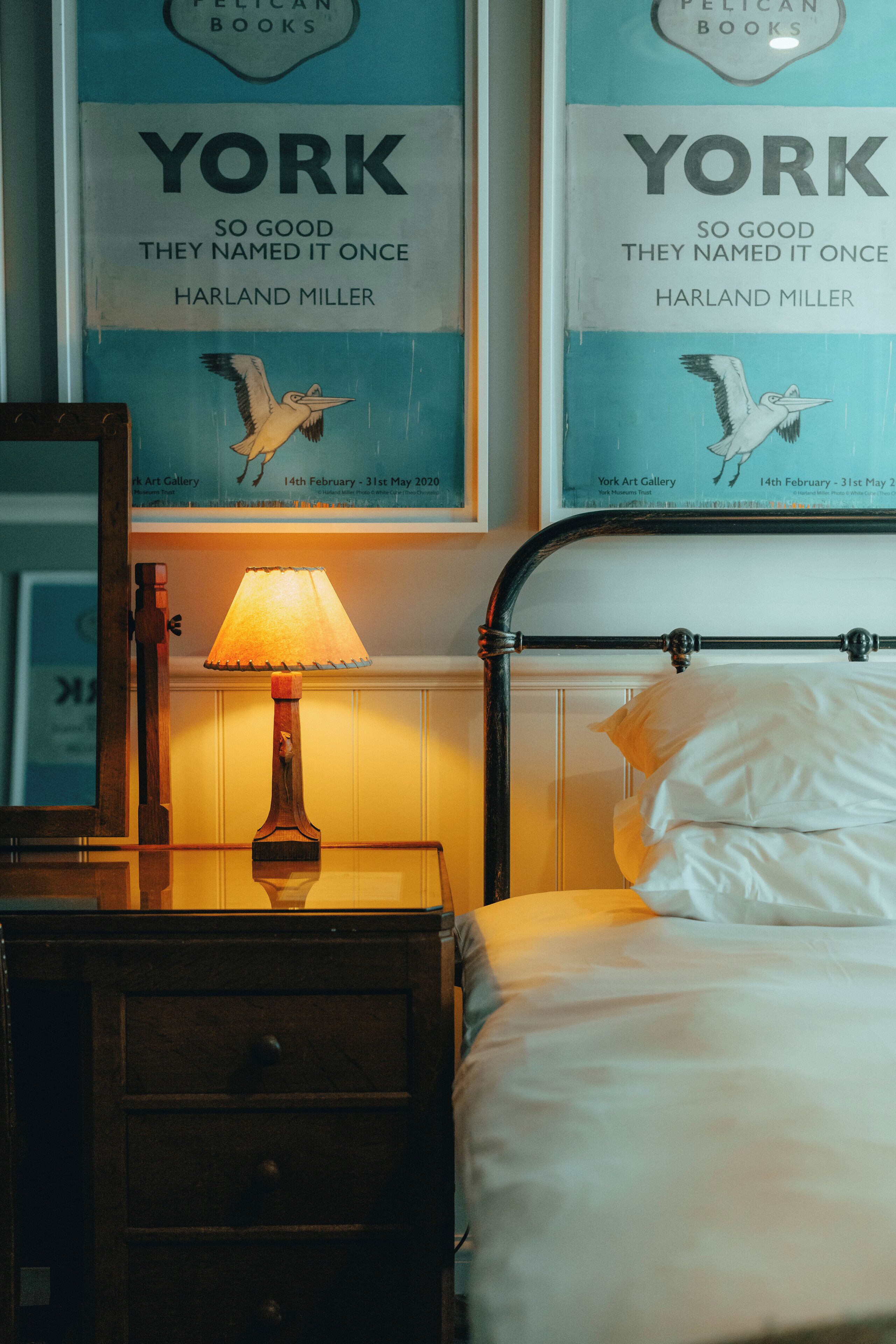 Uma cama bem arrumada com lençóis brancos, uma luminária de cabeceira e dois livros emoldurados intitulados 'York' de Harland Miller na parede acima.