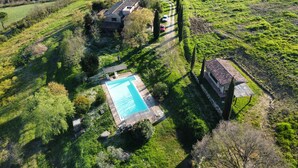 Vista aérea de uma propriedade no campo com piscina, um gazebo e uma pequena casa, tudo cercado por vegetação exuberante e campos.