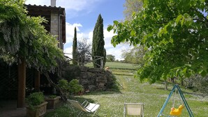 Um jardim com baloiço, cadeira e um balanço. Há plantas em vasos e uma parede de pedra.