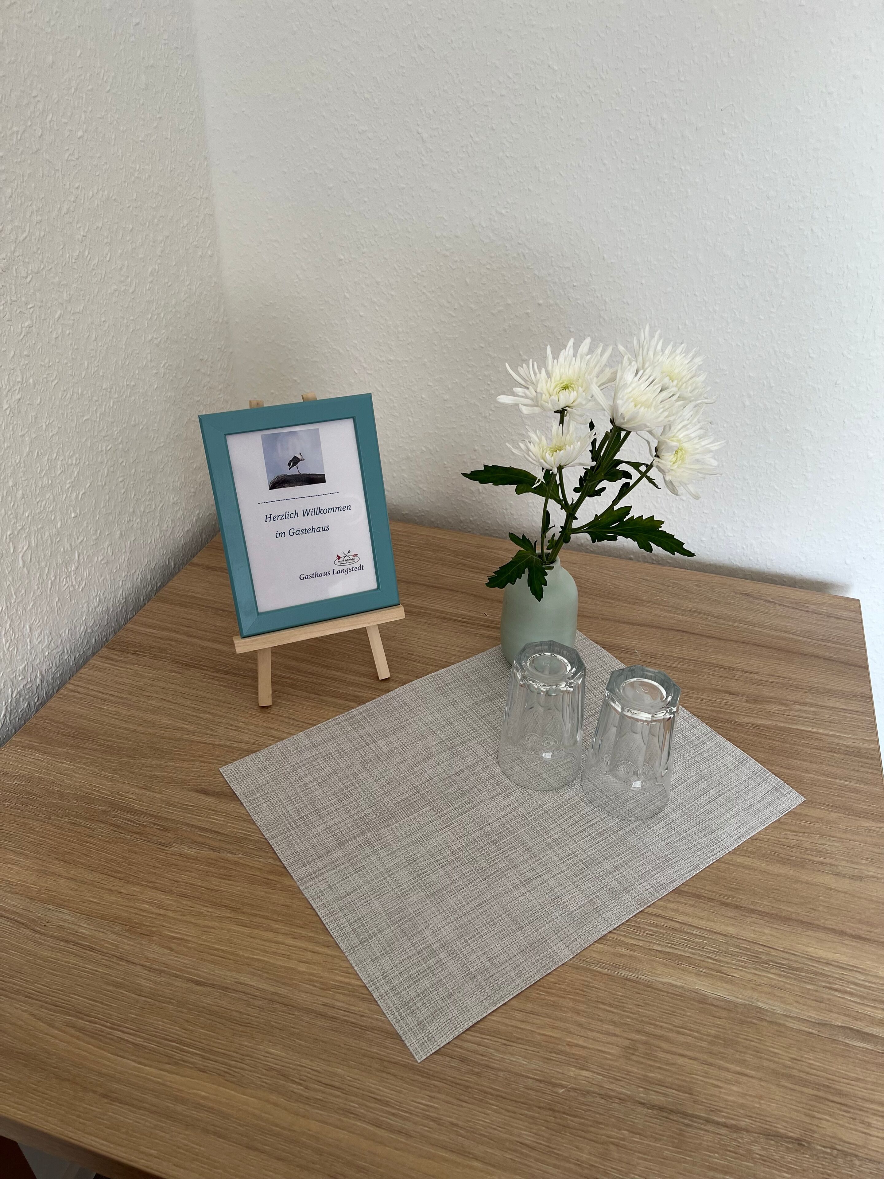 Uma placa de boas-vindas emoldurada, um vaso com flores brancas e três copos de vidro sobre uma mesa de madeira com um jogo americano texturizado.