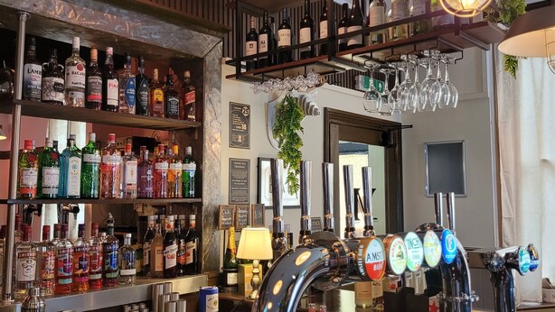 Uma área de bar com diversas prateleiras de garrafas de bebidas, um sistema de torneiras com várias opções de cerveja e um espelho refletindo o interior.