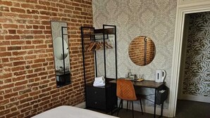 Quarto de hotel com uma cama grande, uma escrivaninha com cadeira, um cabideiro, um espelho redondo e uma parede de tijolos.