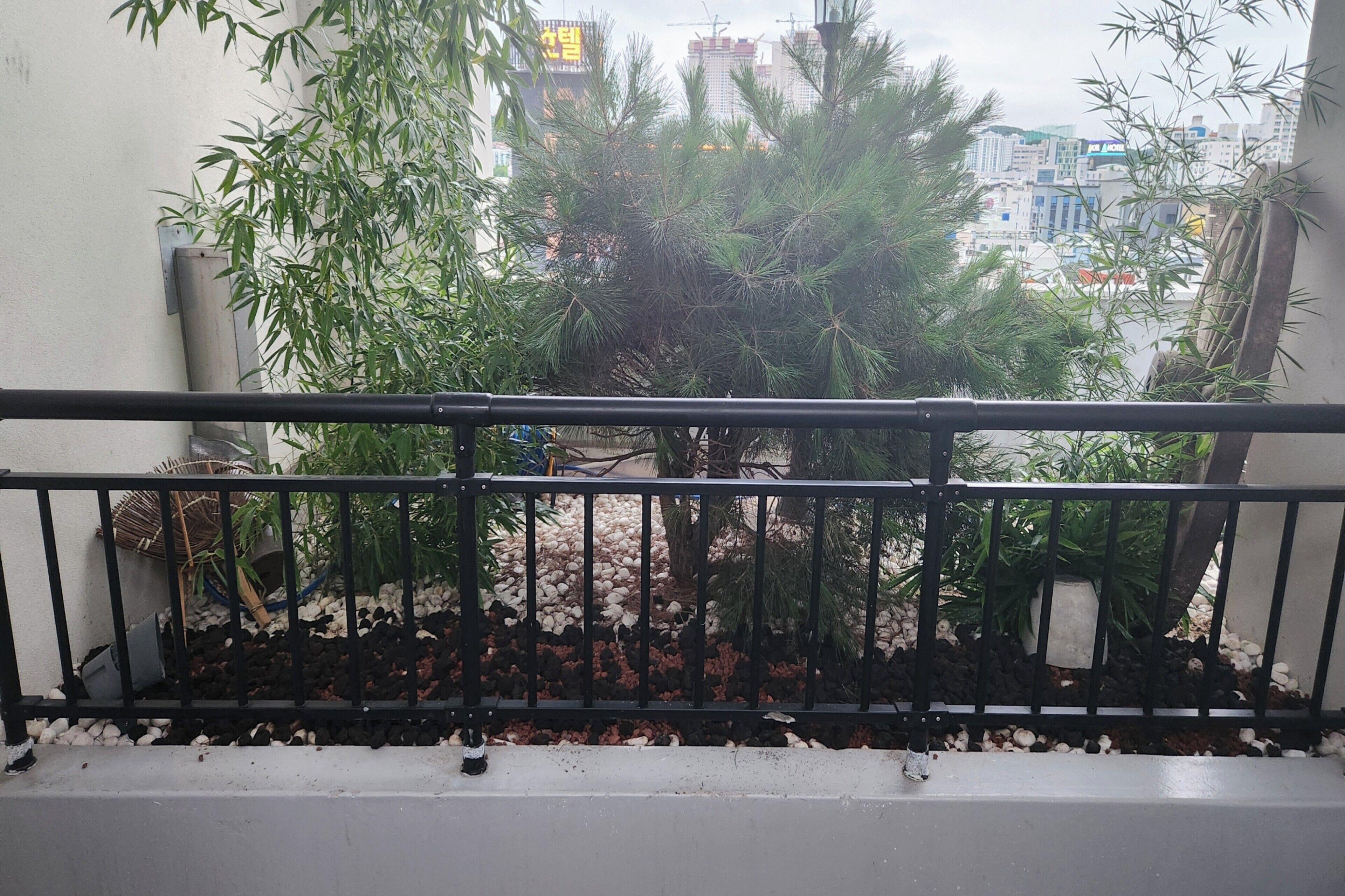 Uma varanda com grades de metal, plantas em vasos e vista para edifícios e guindastes de construção ao fundo.
