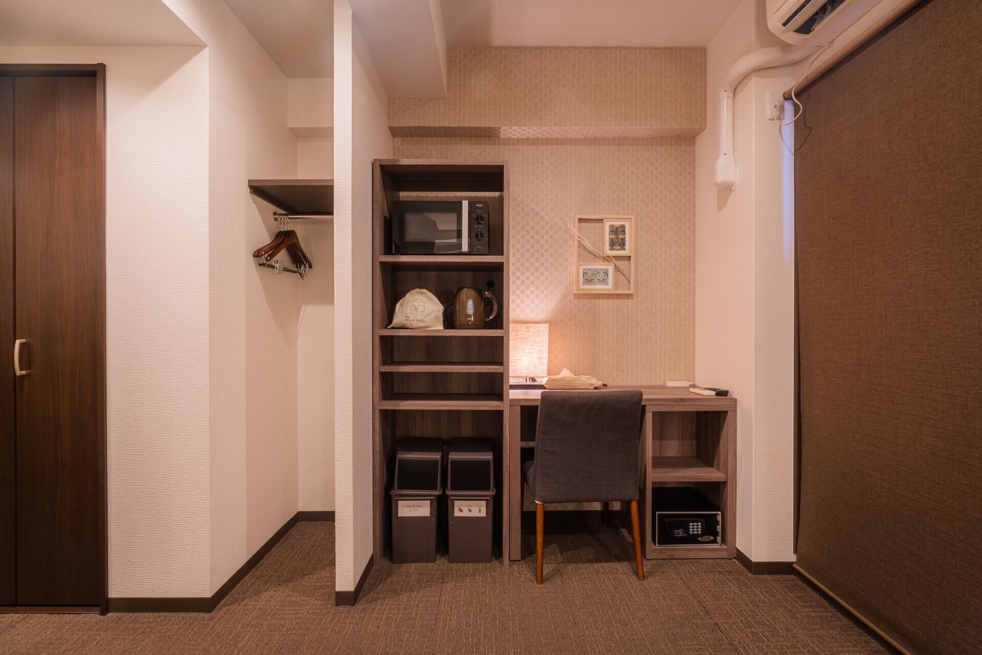 Quarto de hotel compacto com uma secretária de madeira, uma cadeira, micro-ondas, lixeira e pequeno armário.