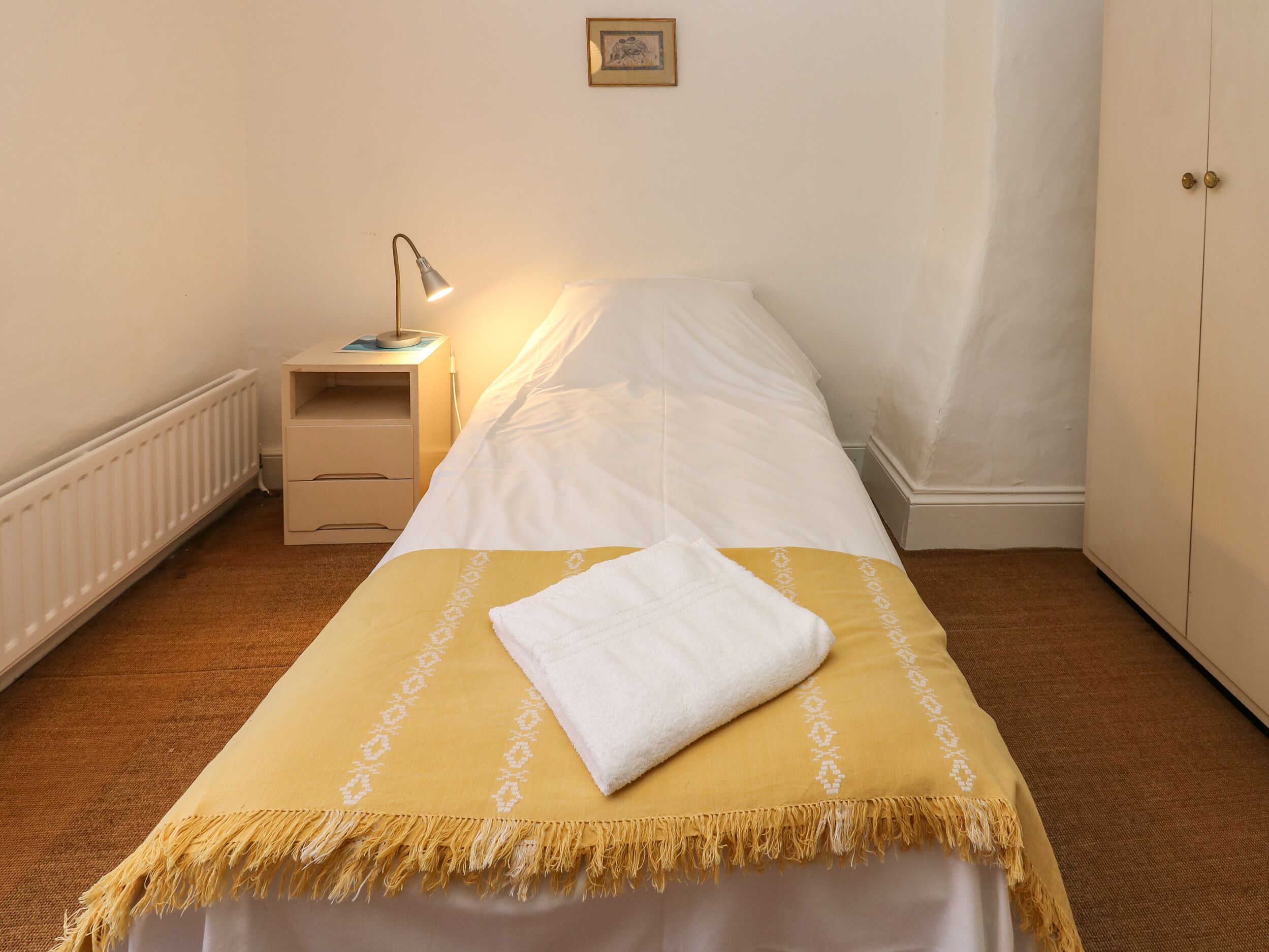 Uma cama bem arrumada com travesseiro branco e manta amarela, luminária de cabeceira, mesa de cabeceira pequena e radiador.