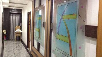 Corredor de hotel com obras de arte emolduradas nas paredes, uma cadeira de madeira e um vaso com flores.