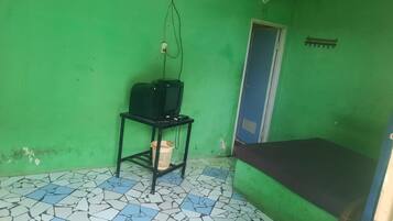Quarto com parede verde, chão de azulejos, uma mesinha com televisão, porta e cama.