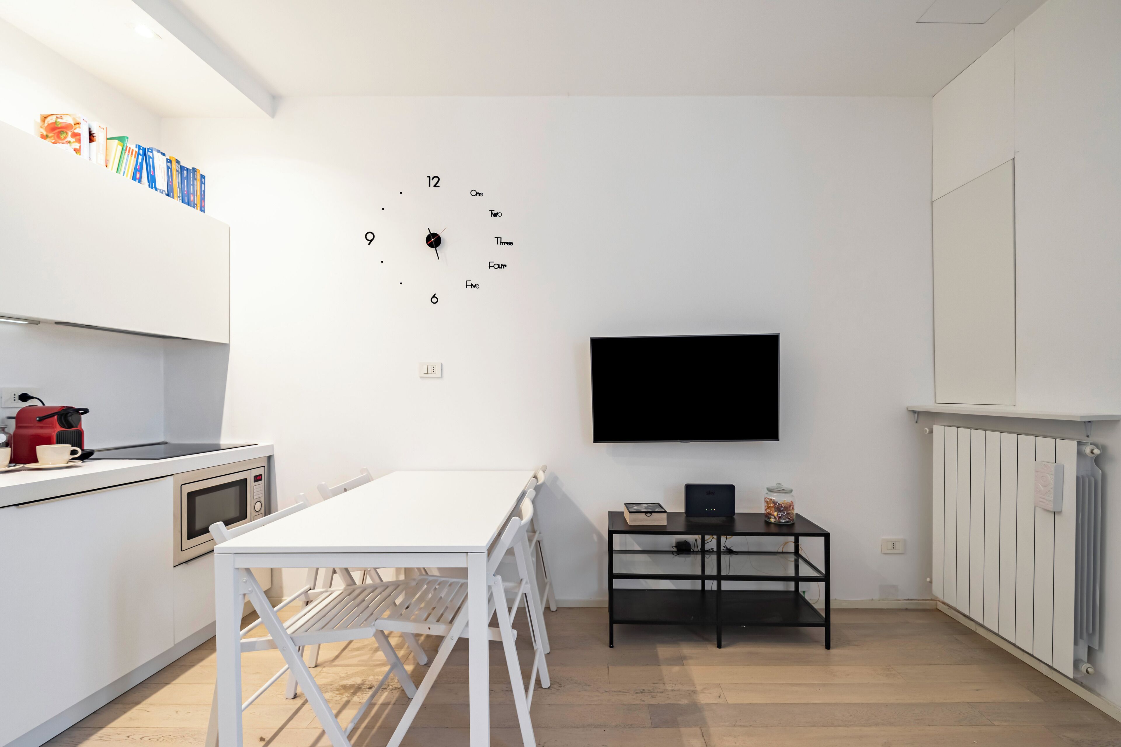 Cozinha moderna com bancada branca, micro-ondas embutido e chaleira vermelha. Uma mesa de jantar branca com cadeiras está posicionada em frente à bancada. Uma televisão de tela plana está montada na parede acima de uma mesinha lateral preta com um suporte e uma pequena planta.
