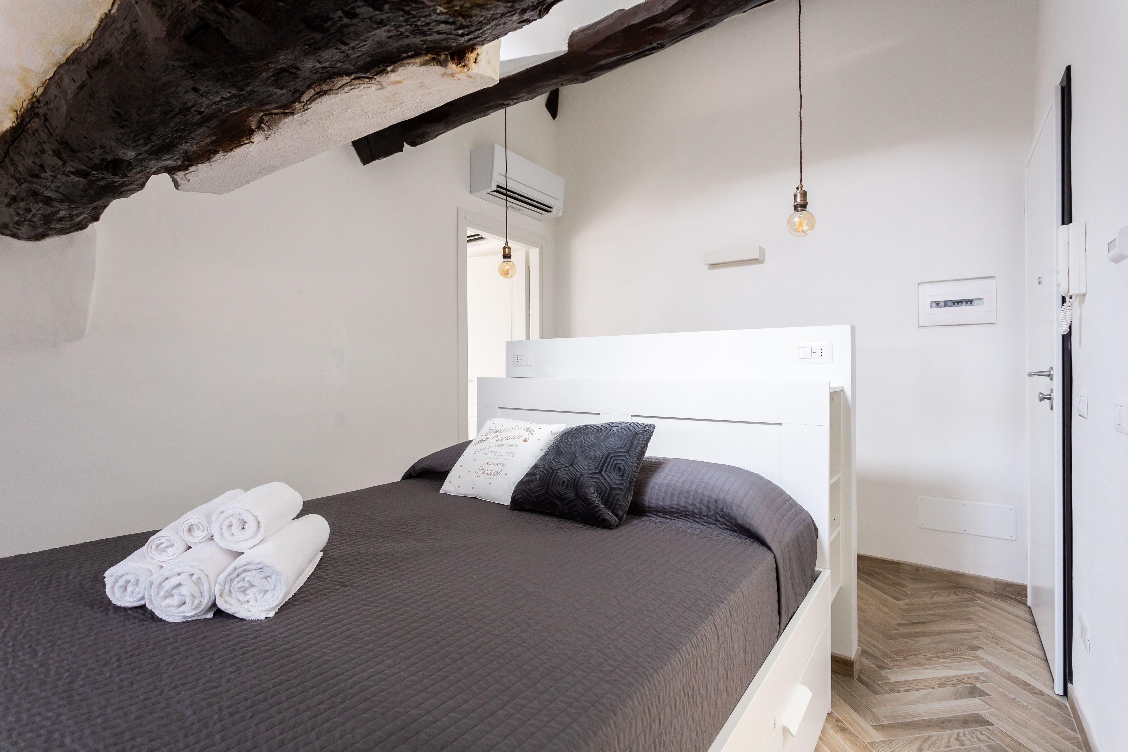 Cama bem arrumada com toalhas enroladas e um travesseiro, cabeceira branca e piso de madeira.
