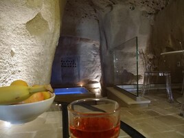 Interior com paredes de pedra, mesa de vidro, uma tigela de frutas e um copo de bebida vermelha.