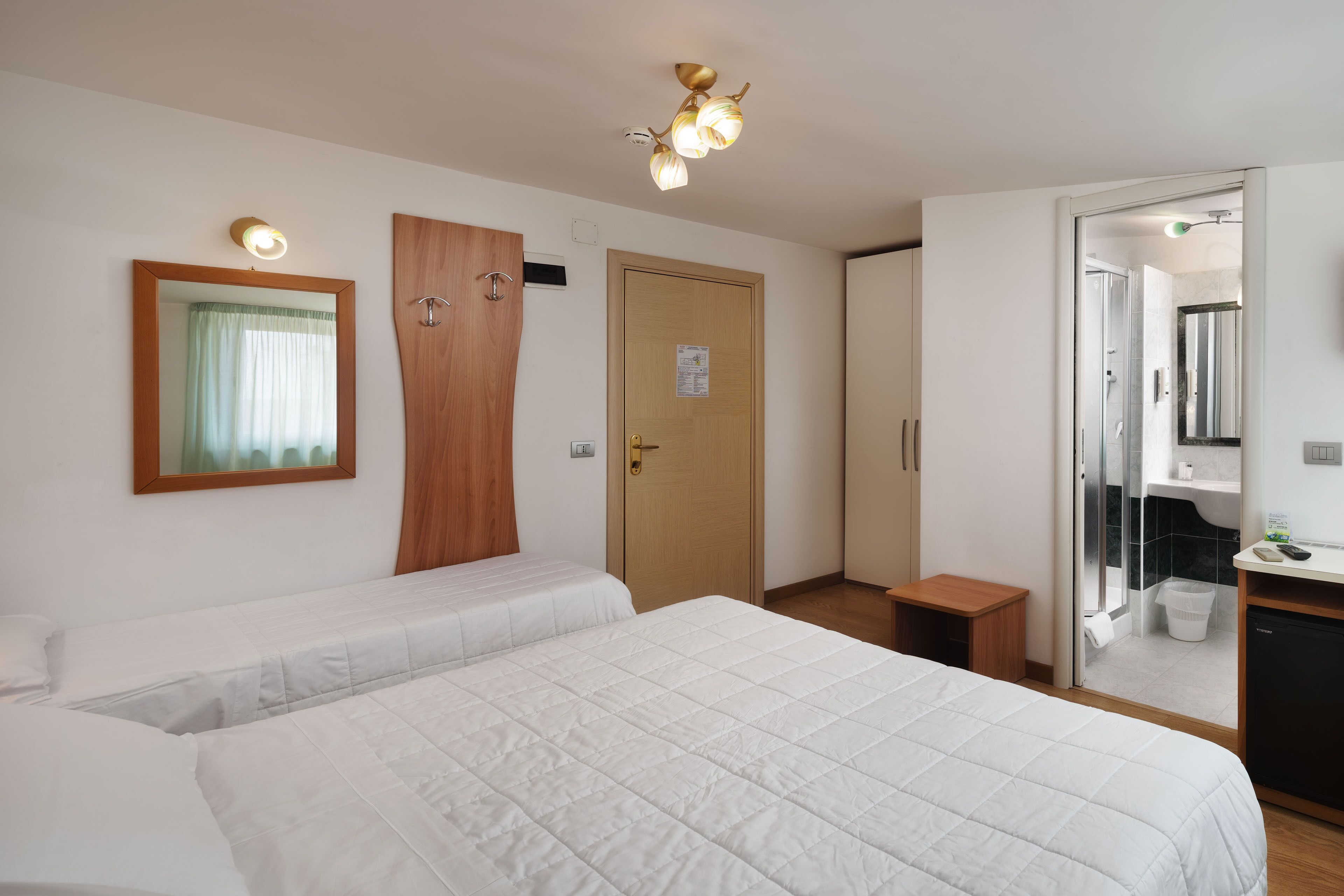 Quarto de hotel com uma cama grande, uma porta de madeira, uma mesinha de cabeceira pequena e um banheiro visível através de uma porta aberta.