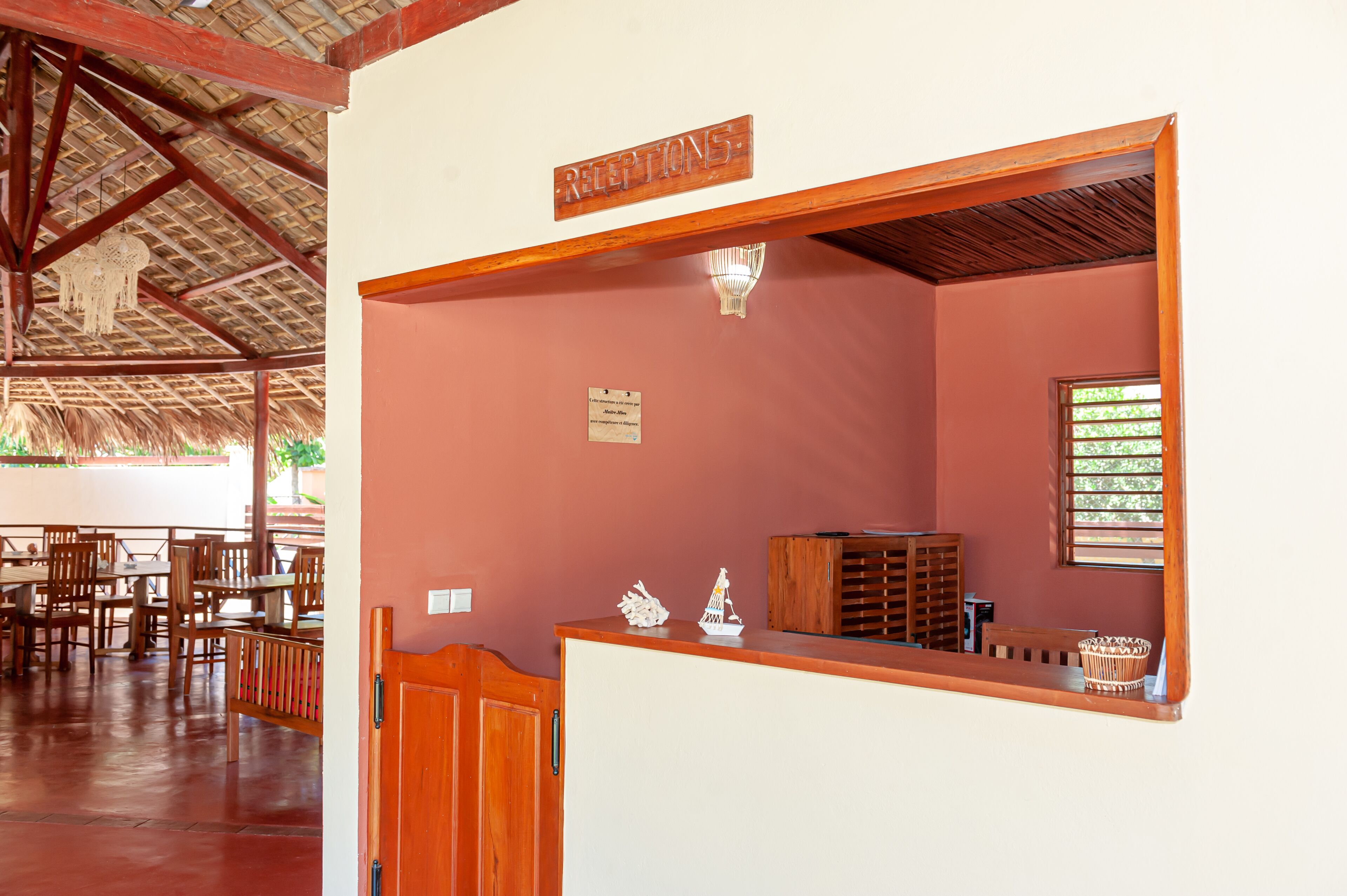 Uma área de recepção com balcão de madeira, uma placa acima e vista para a sala de jantar com mesas e cadeiras.