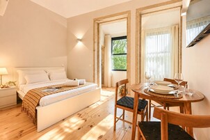 Quarto com cama, mesa de cabeceira, luminária e um espaço de jantar com mesa, cadeiras e janela.