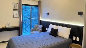 Quarto moderno com uma cama grande, uma mesinha redonda, uma cadeira e vista para o exterior através de uma janela.