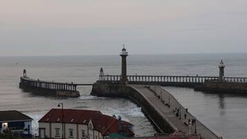 Um cais costeiro com um farol, um muro de pedra e uma fileira de edifícios com telhados vermelhos.