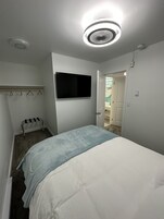 Quarto com cama, TV fixada na parede, cabideiro e porta que dá para outro cômodo.